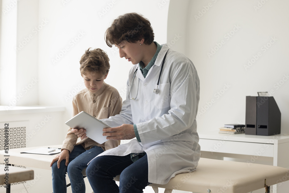 Engaged young pediatrician man showing content on tablet to patient kid ...