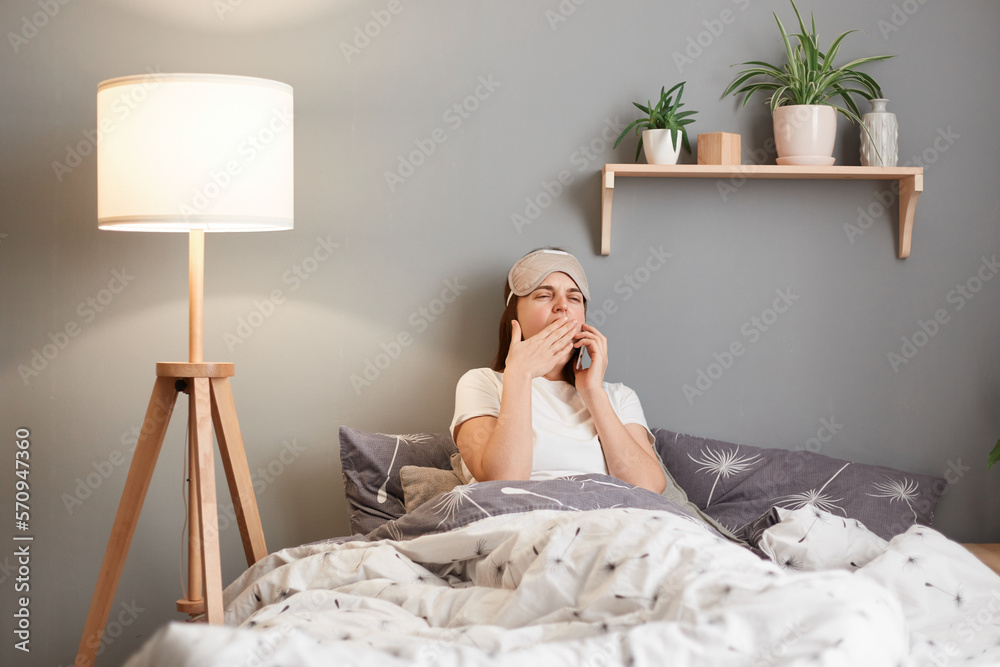 Foto de Portrait of sleepy young adult woman in bed yawning and talking on phone, wearing ...