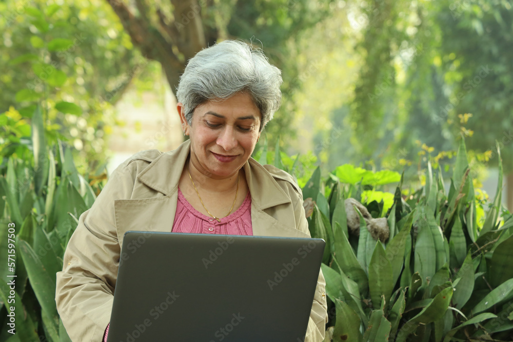 Happy Indian woman working on a Laptop in a lush green park in day time ...