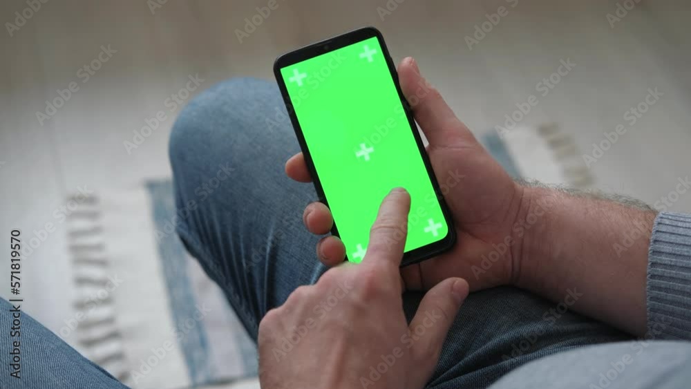 Young man sitting at home holding smartphone green mock-up screen in ...