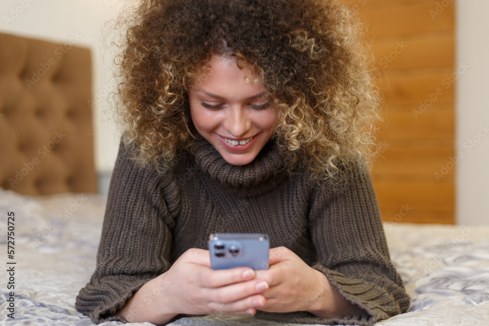 Happy white woman with curls and diastema lying on bed and texting with a smart phone. Portrait ...
