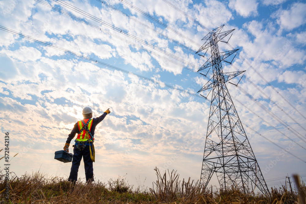 A technician stands holding a toolbox while pointing at a high-voltage ...