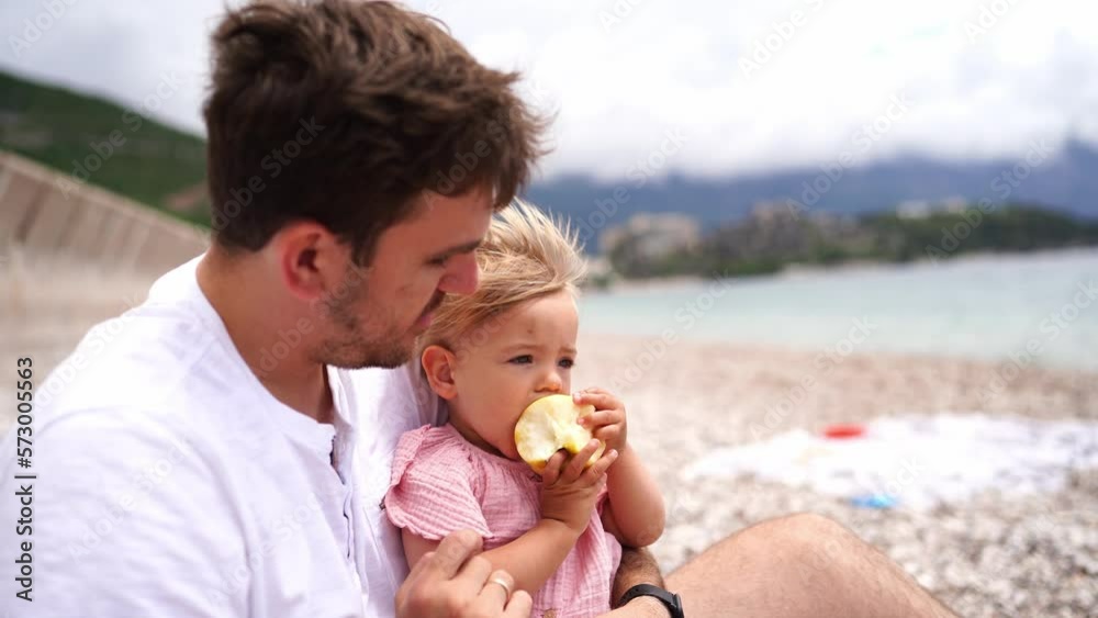Dad pats the head of a little girl eating an apple while sitting on his ...