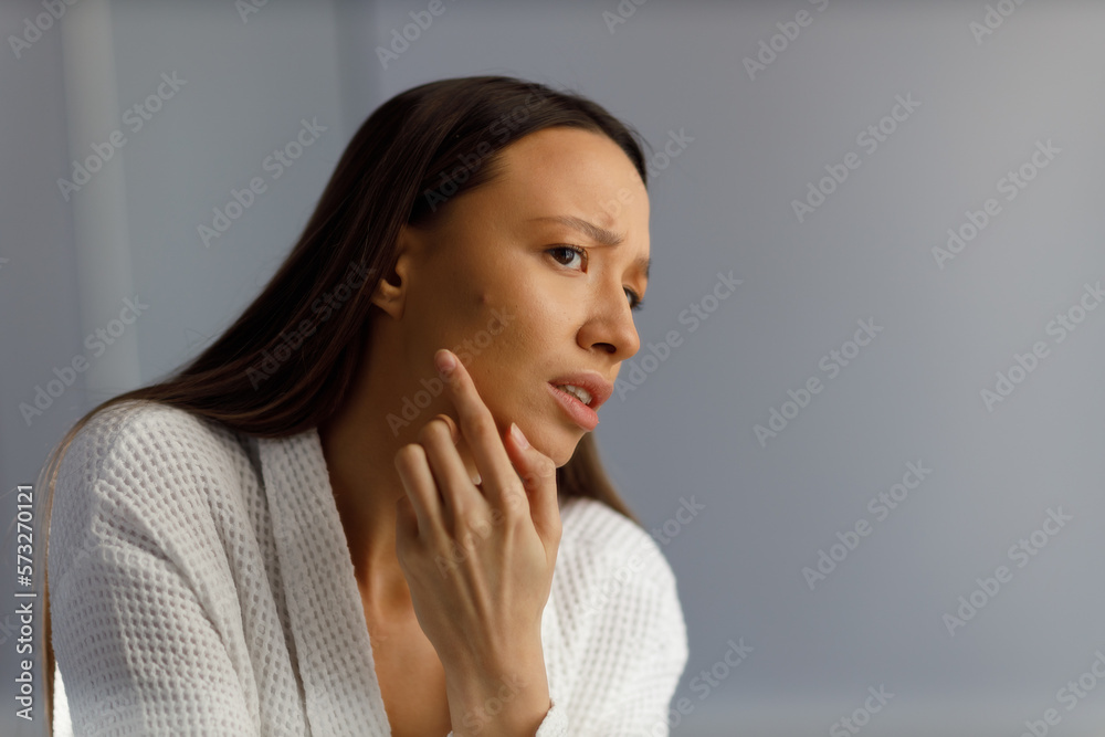 Attractive upset young woman in white bathrobe examines pimples on her ...