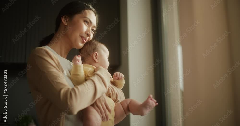 Beautiful Young Asian Mother Holding her Baby in her Arms While Standing Next to a Window at ...