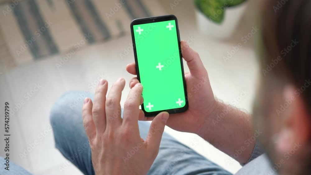 Young man sitting at home holding smartphone green mock-up screen in hand. Male person using ...