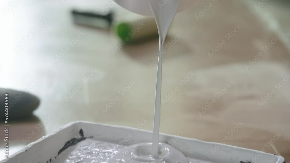 The hands of an unrecognizable person pour white paint from a bucket ...