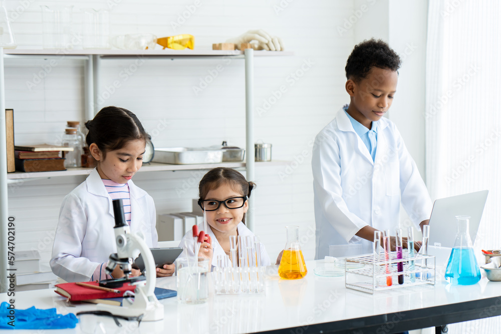 Two little Asian girls and one young African boy in white lab coat with many laboratory tools on ...
