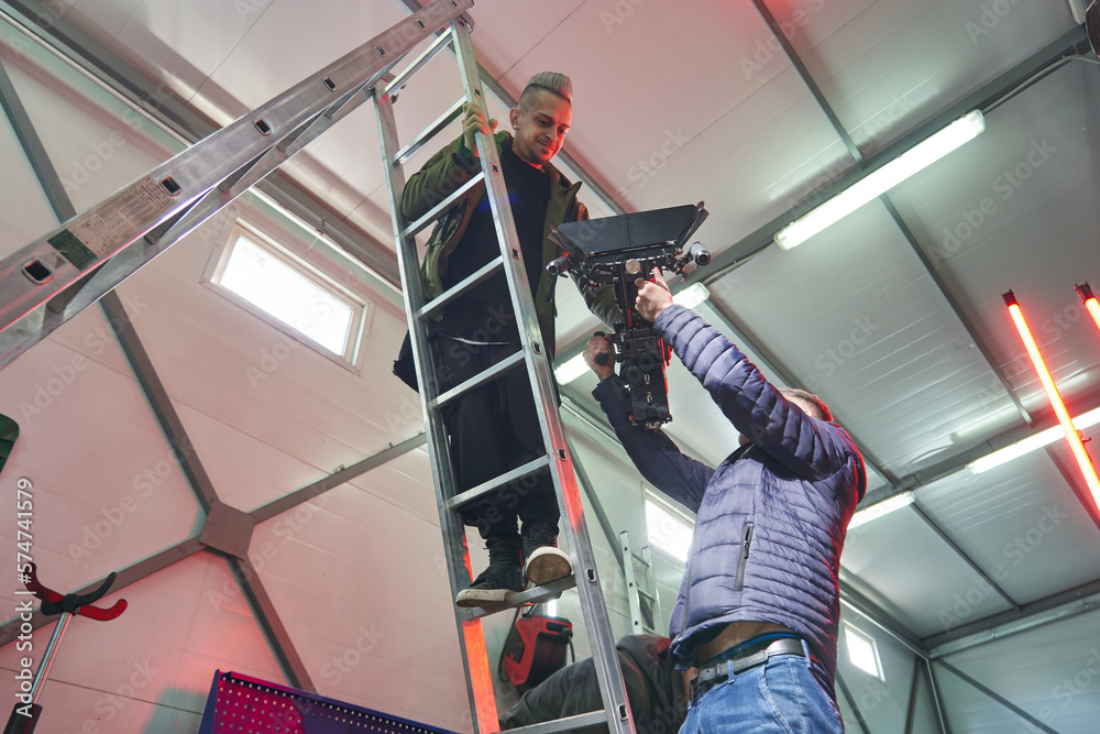 two caucasian men lift a professional movie camera on a high ladder ...