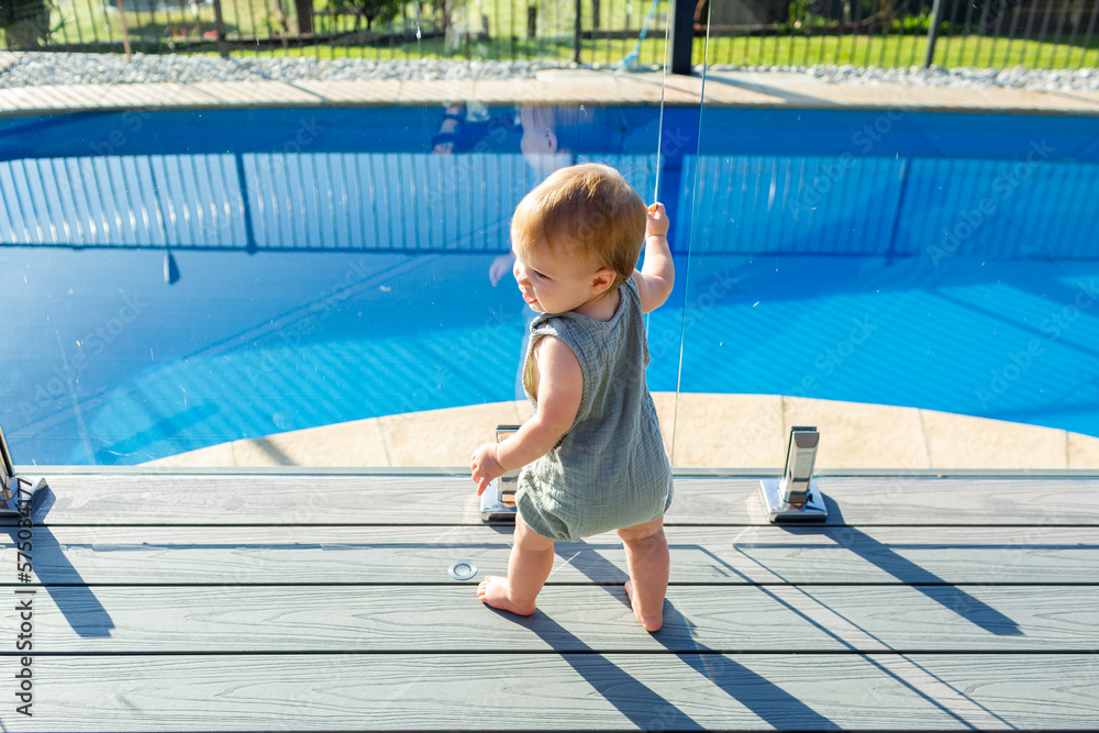 Toddler baby ten months old standing at glass pool fence beside ...