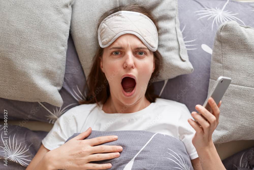 Portrait of shocked surprised young woman wearing sleeping mask lying ...