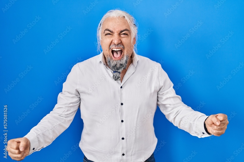 Middle age man with grey hair standing over blue background angry and ...