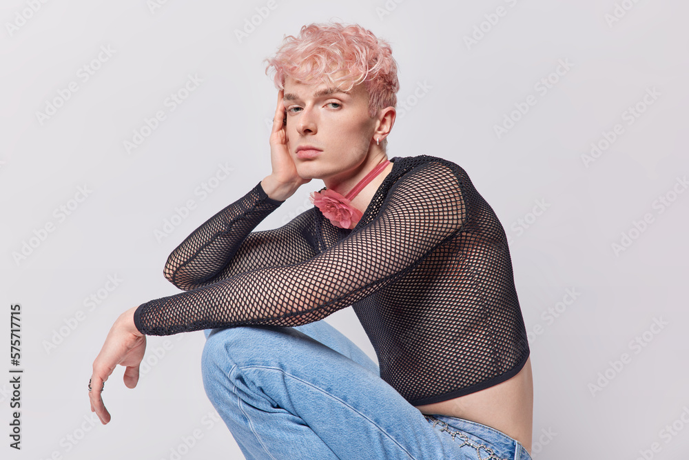 Non-binary person keeps hand on face looks directly at camera squats against white background ...