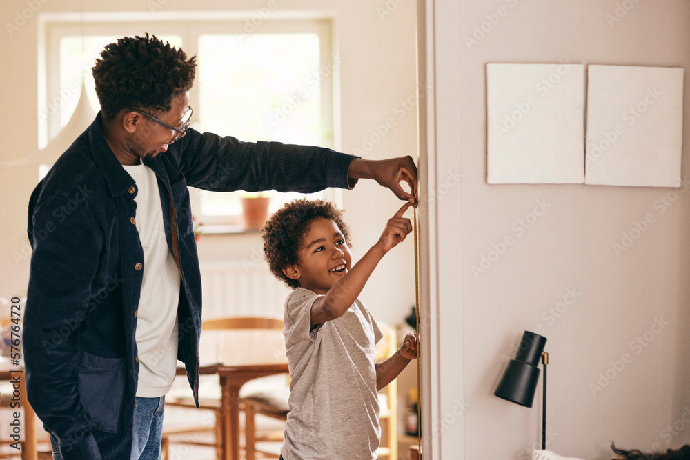 Happy father and son checking height while standing near wall at home ...