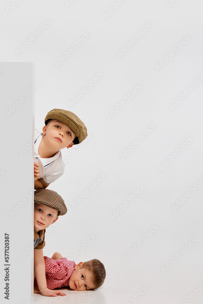 Three little kids, boys peeking out corner, playing against grey studio ...