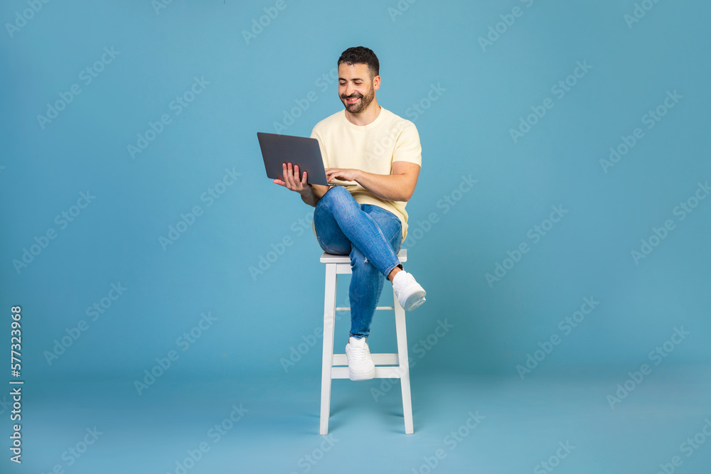 Happy man using laptop computer sitting on chair, working online over blue studio background ...