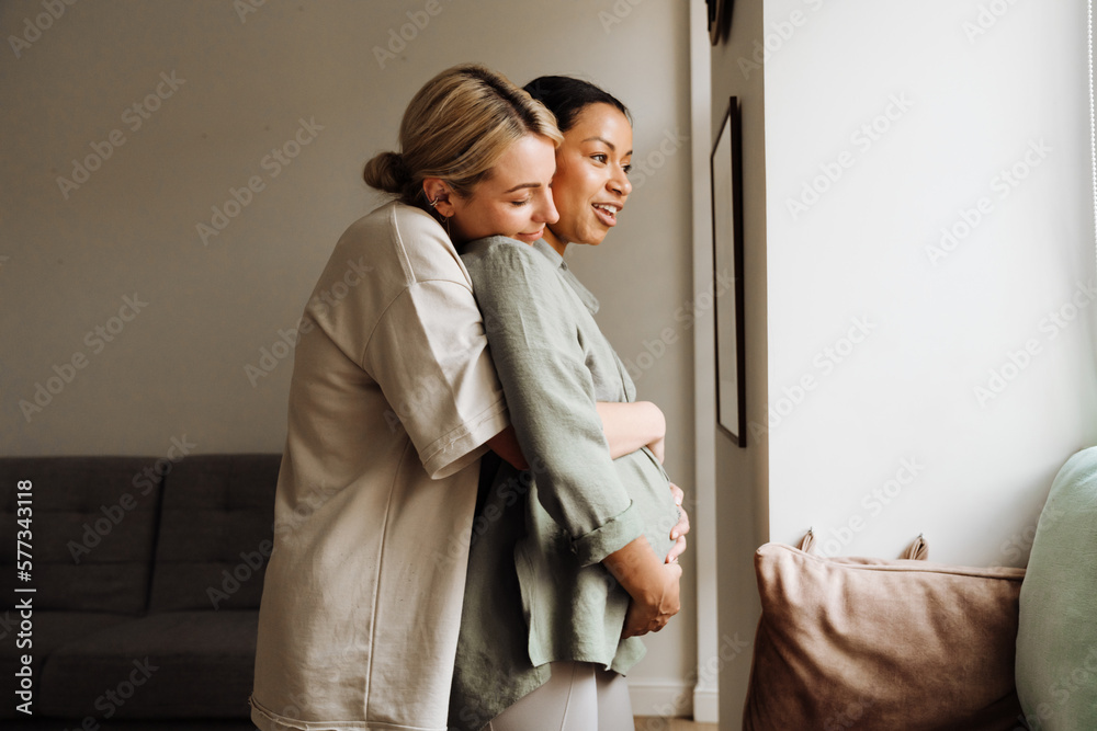 Pregnant lesbian couple hugging and looking through window while standing together at home Stock ...