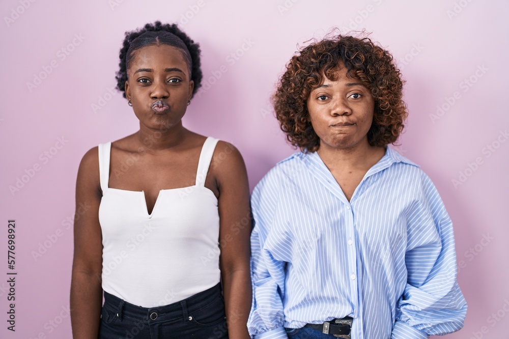 Two african women standing over pink background puffing cheeks with ...