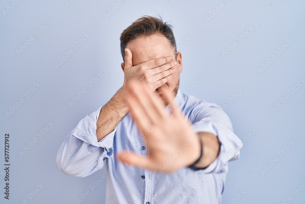 Middle age caucasian man standing over blue background covering eyes ...