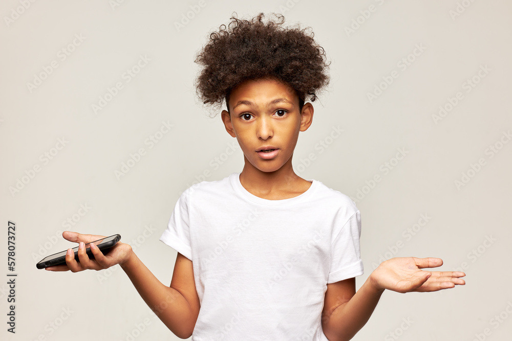 Studio image of puzzled upset confused kid boy shrugging with mobile ...