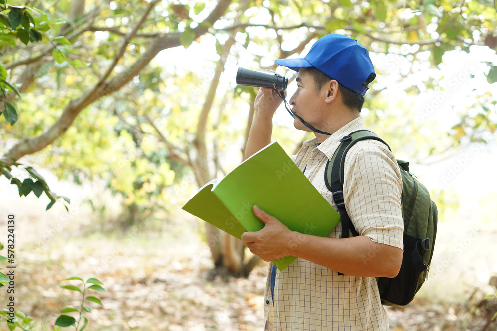 Asian man explorer wears blue cap, holds binocular in forest to survey ...