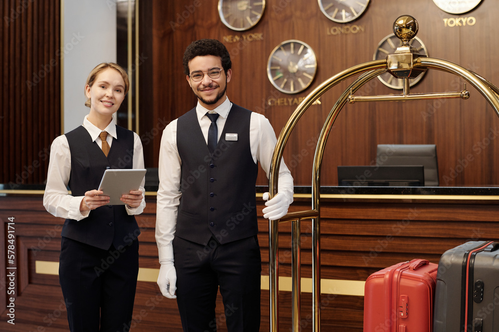 Happy young elegant receptionist and bellboy standing in lounge of luxurious hotel and looking ...