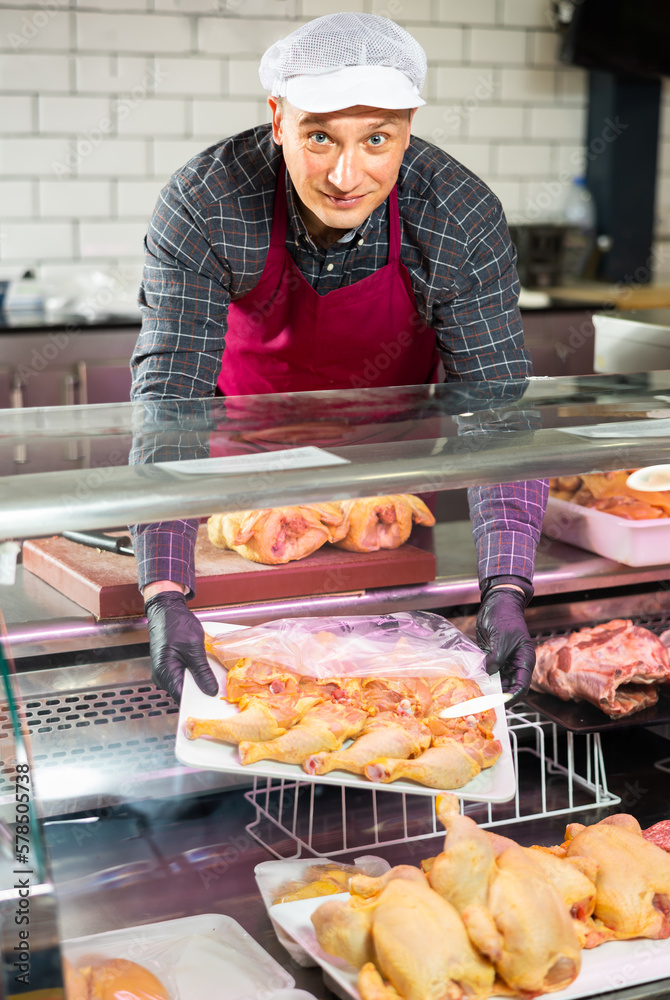 Smiling adult butcher shop worker laying out fresh raw produce in glass ...