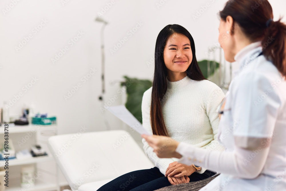 A smiling Asian female patient talking with a female doctor, sitting in ...