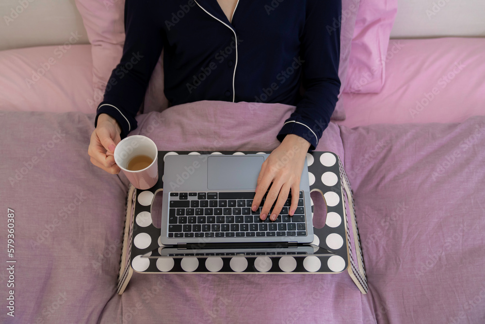 woman teleworking at home with laptop lying in bed drinking tea wearing ...