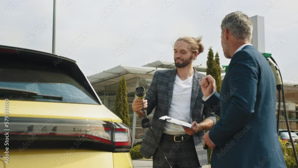 Two men near car charging station. Young confident salesman showing ...