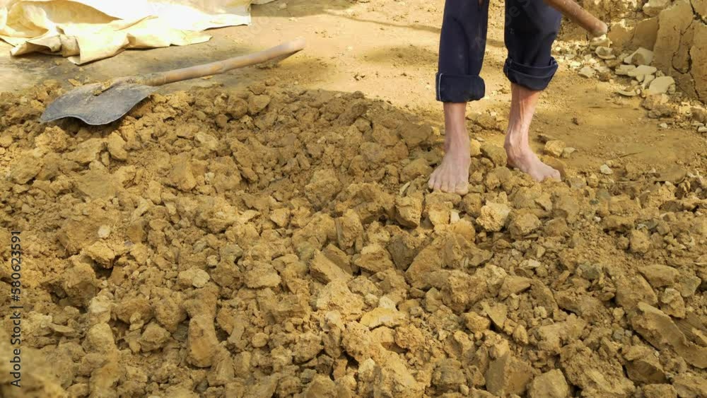 The sun shines on a barefoot man breaking clay soil, showing the hard ...