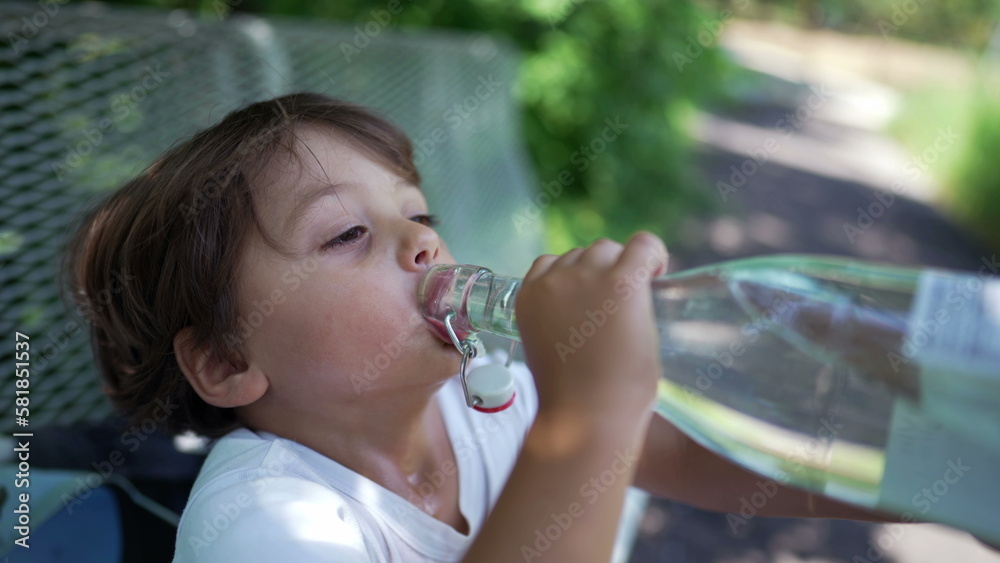 Foto de Child drinks water during hot summer day. Little boy gulping from glass bottle hydrating ...