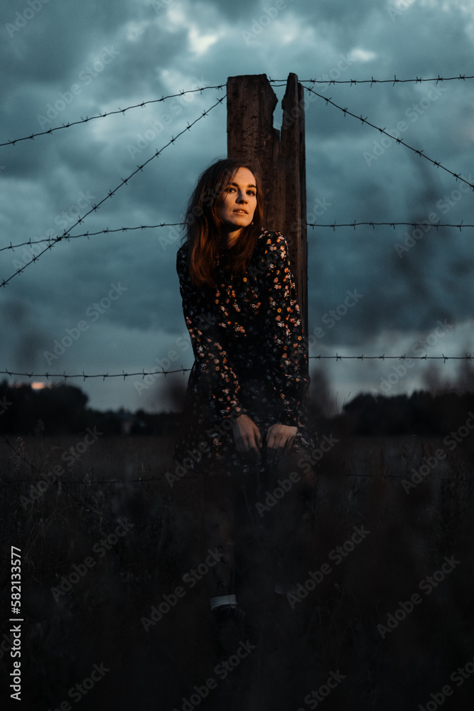 A girl in a dress stands in a field at night surrounded by barbed wire ...