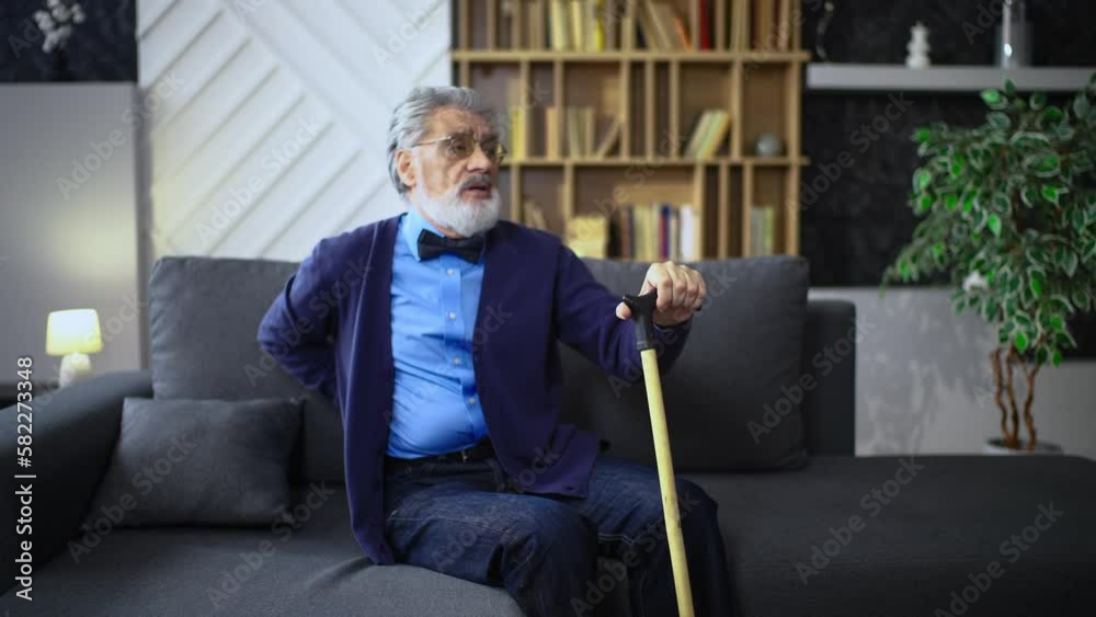 Senior man in glasses sits with a cane, got up from the sofa, his back ...