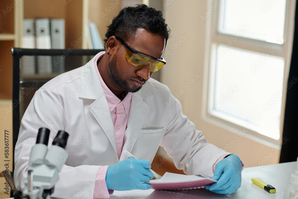 Foto de Young man in lab coat and gloves making notes in copybook while ...