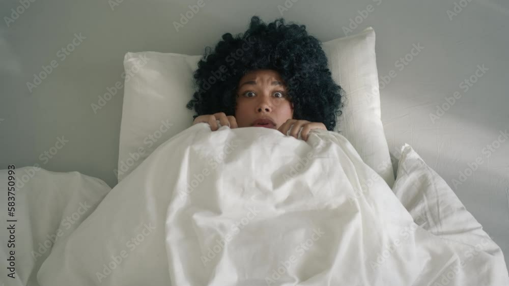 Scared african american woman peeking out from her blanket in bed with ...