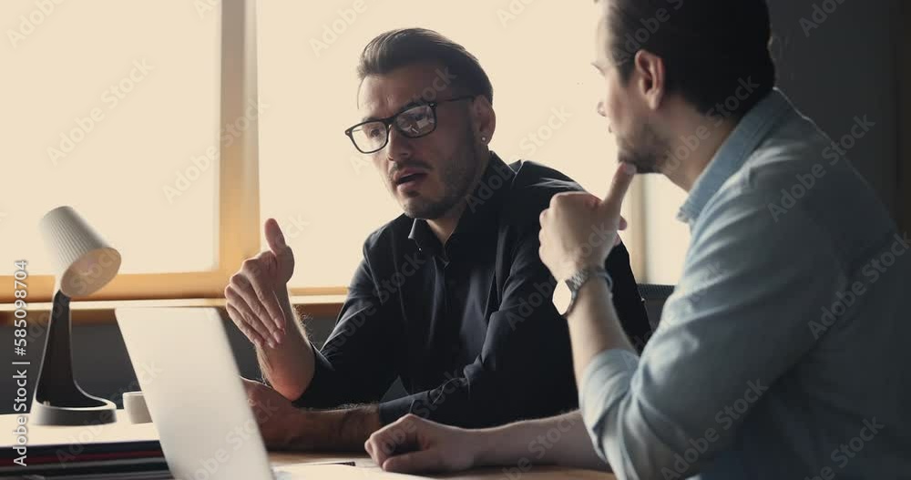 Two serious millennial employees talking at laptop, sitting at ...