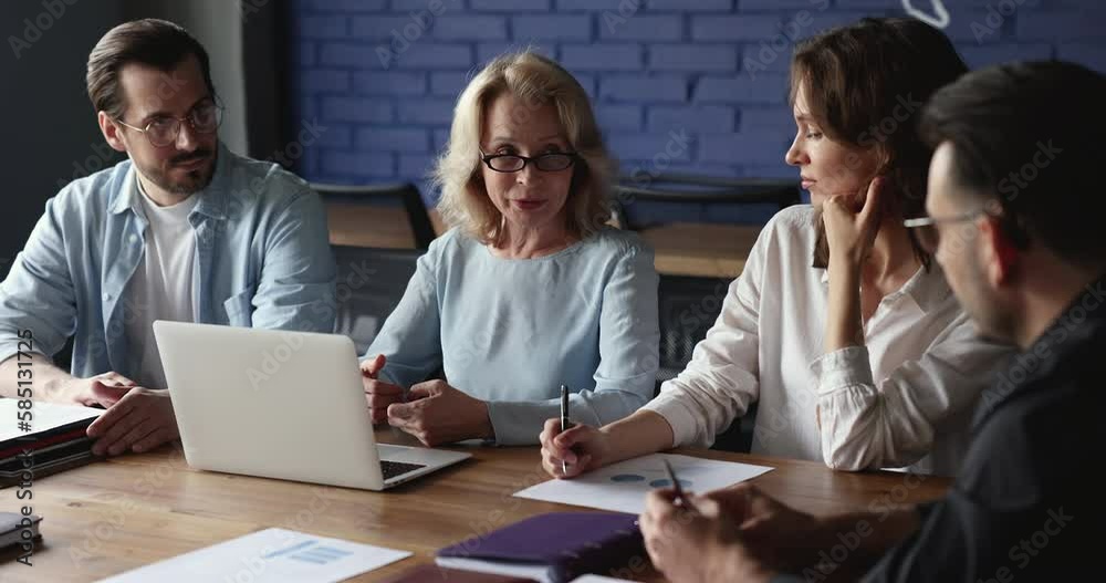 Senior mentor woman sitting at table with laptop, talking to team of ...