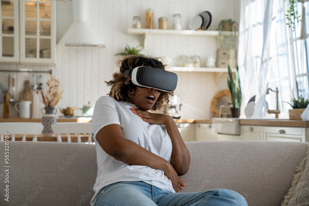 Shocked young black woman sitting on sofa in virtual reality headset ...