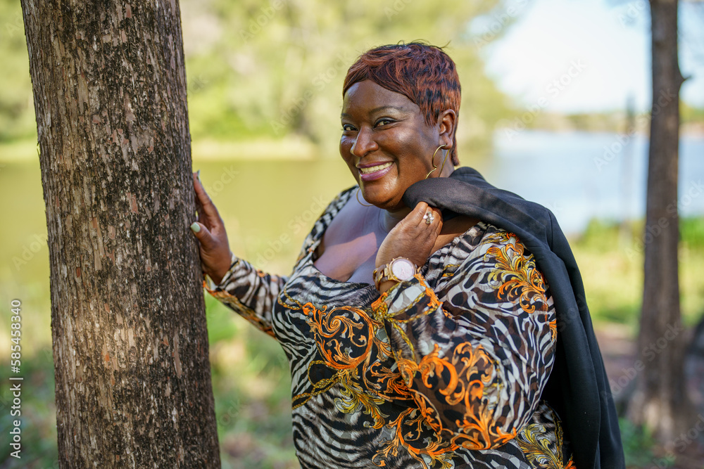 Beautiful black plus sized model posing by a tree in a tranquil park ...