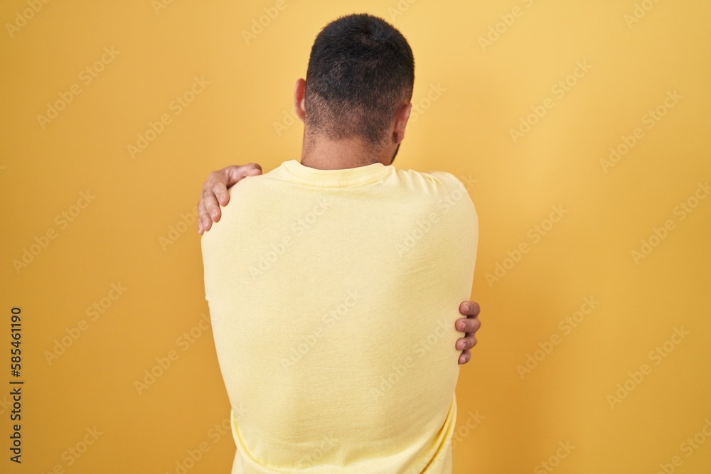 Young hispanic man standing over yellow background hugging oneself ...