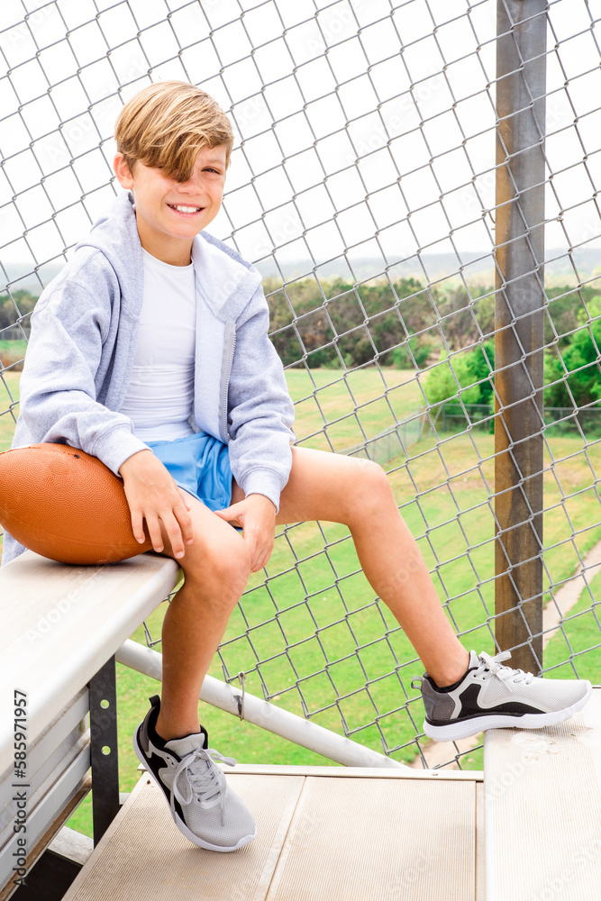 Young preteen boy sitting on bleachers with football outside in the sun ...