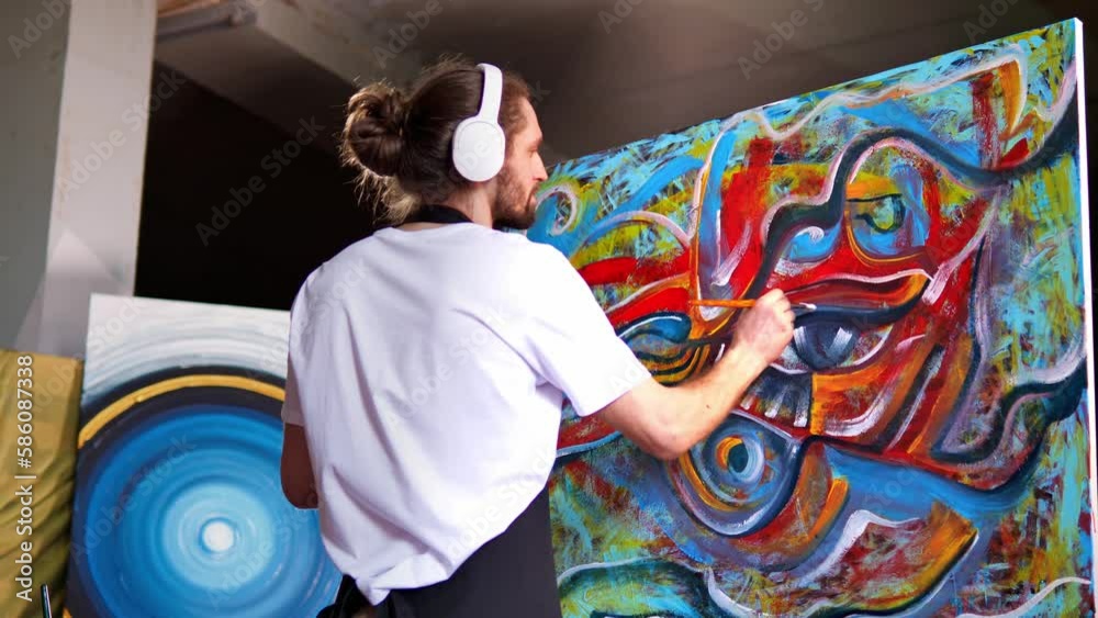 Talented innovative artist in white headphones paints with brushes on a large canvas in studio ...
