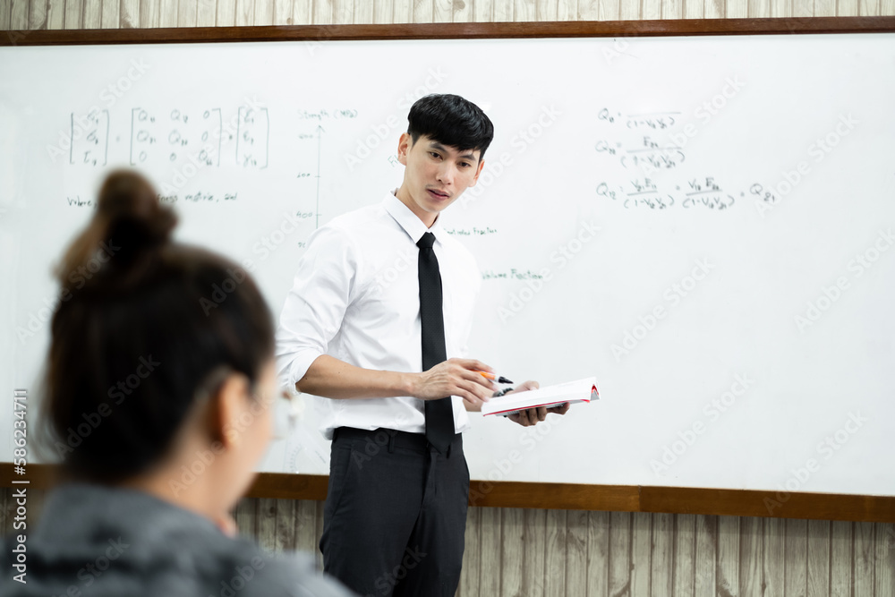 An Asian male teacher in a classroom near the whiteboard is writing and ...