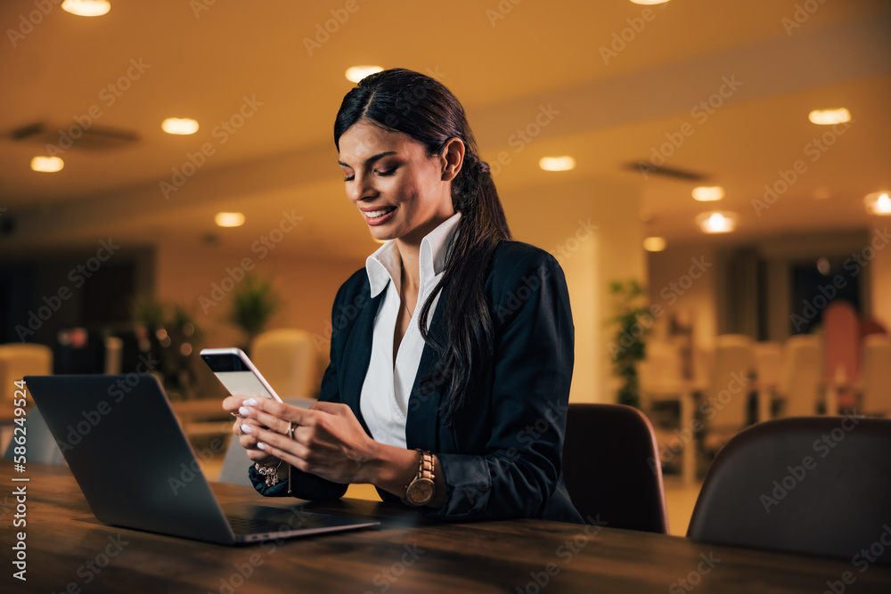 Happy businesswoman sitting in front of a laptop, using a mobile phone ...