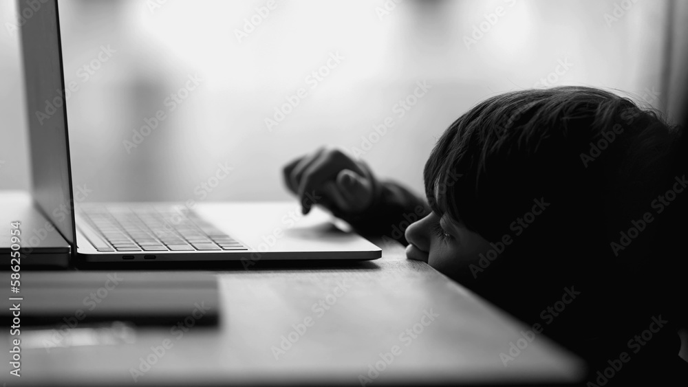 One bored little boy leaning on table and using computer laptop feeling ...