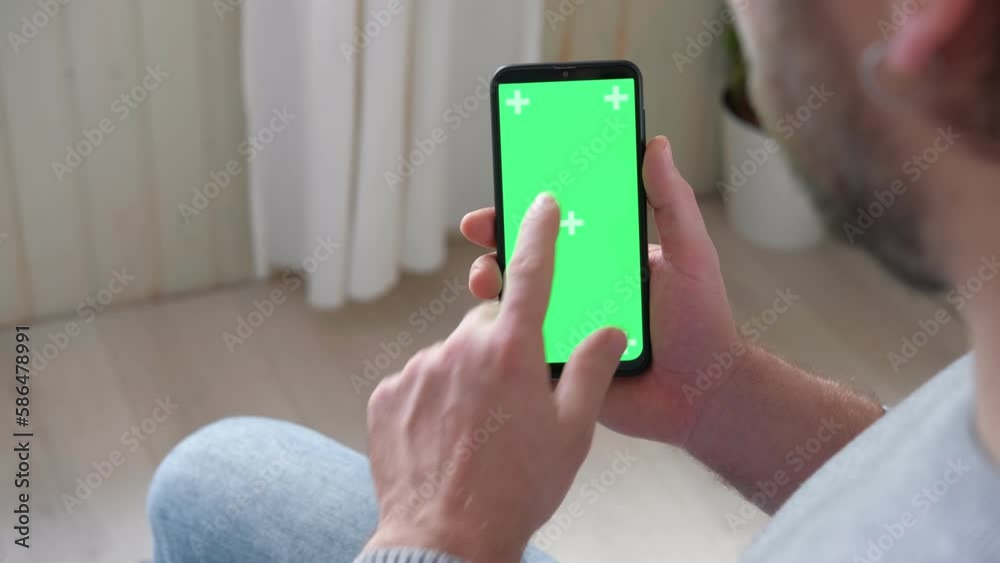 Young Guy sitting at home holding smartphone green mock-up screen in hand. Male person using ...