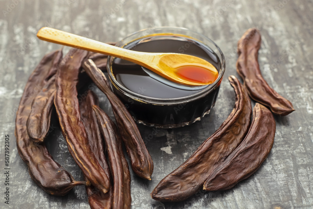 carob molasses in glass bowl and in wooden spoon and carob pods on ...