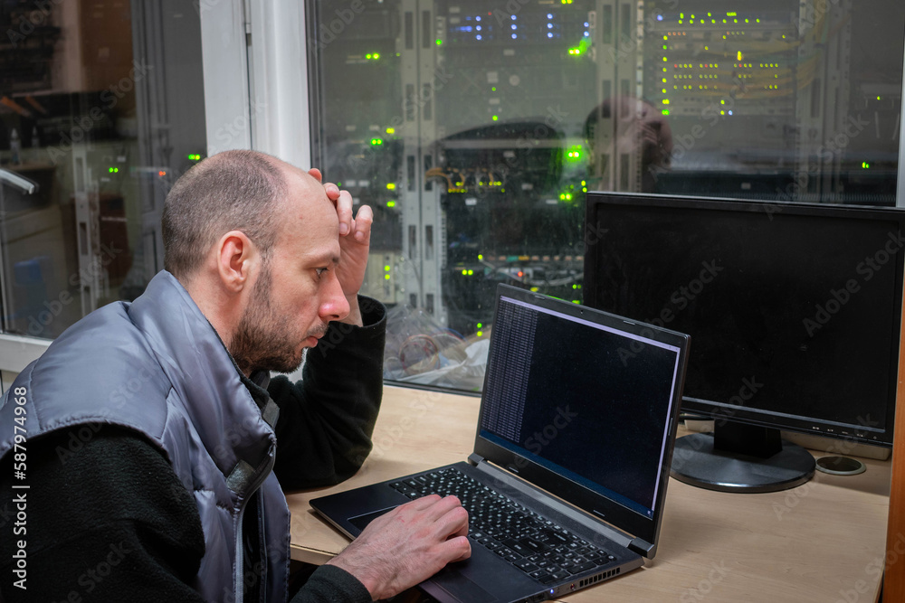 A man works on a laptop next to a server room. A programmer sits at a ...