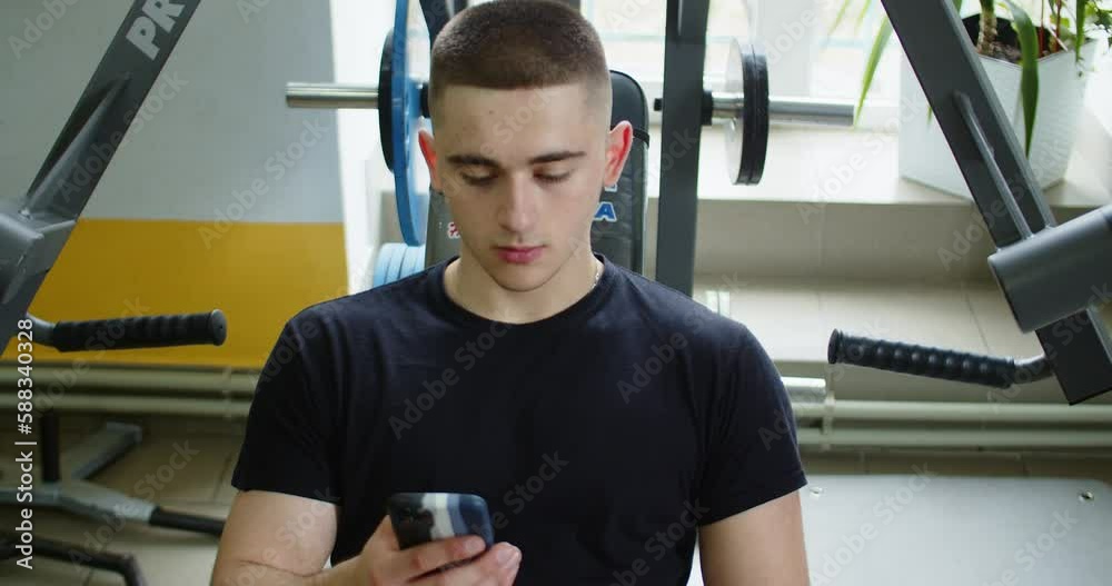 Portrait of guy in gym while resting between a workout uses phone body ...